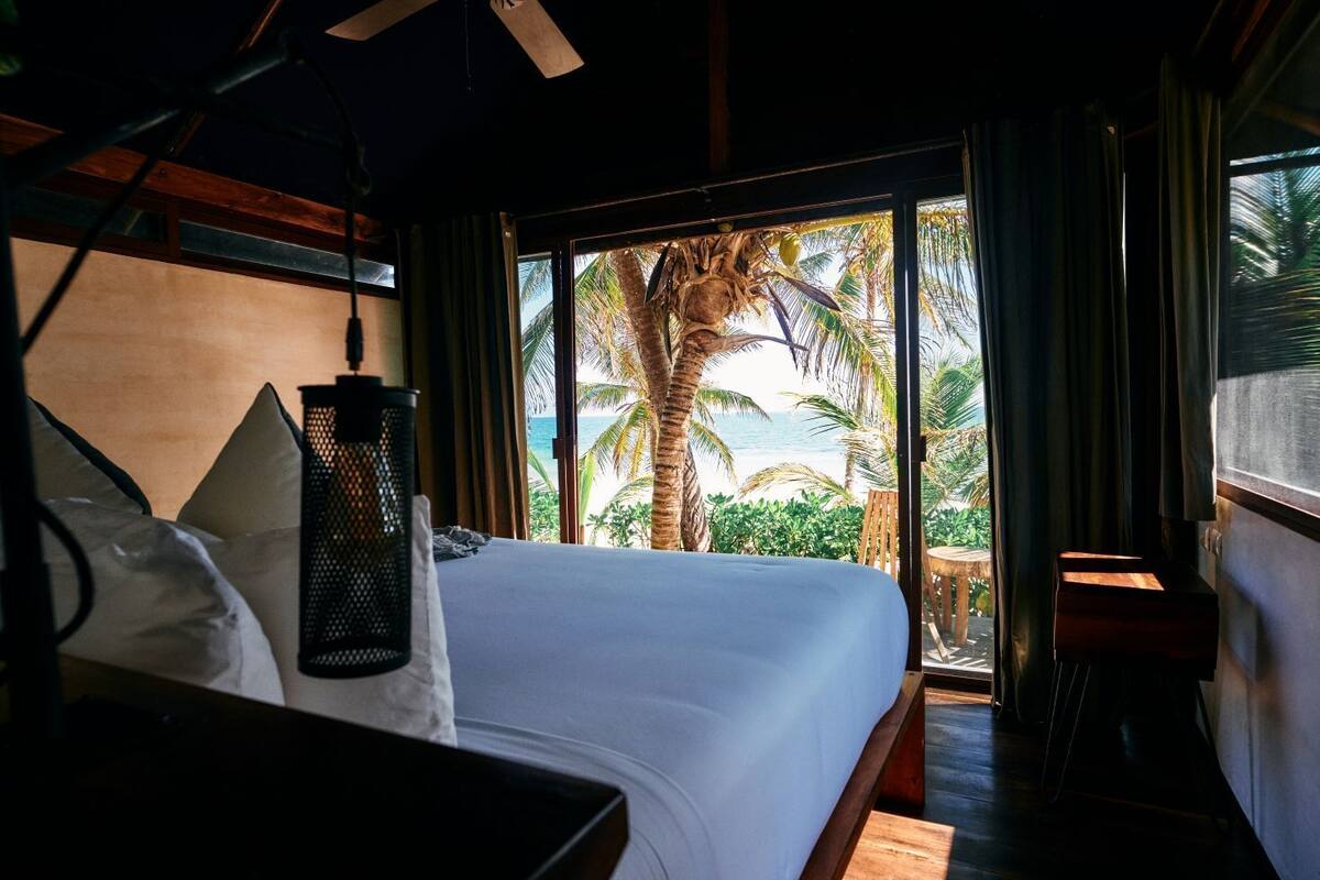 Quarto com cama grande, ventoinha no teto e vista para a praia e palmeiras através de uma janela ampla.