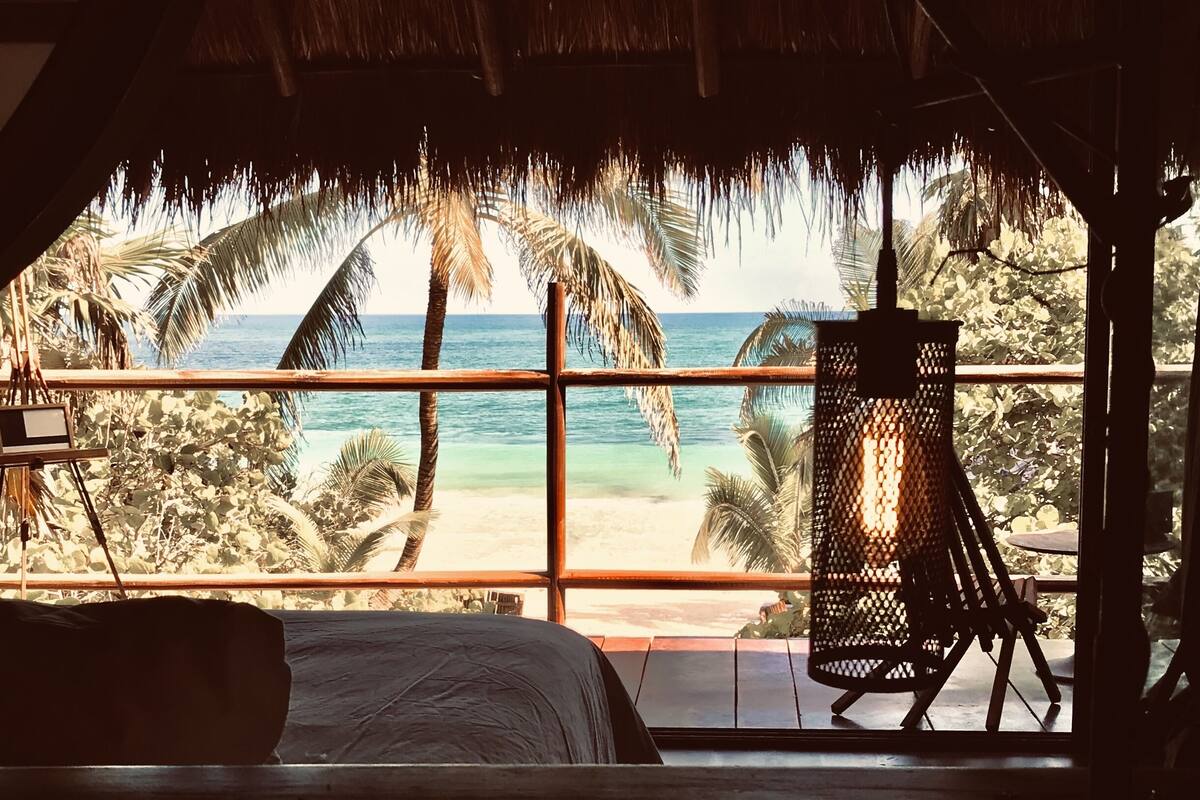 Quarto com uma grande janela de onde se vê a praia, uma cama e uma cadeira suspensa.