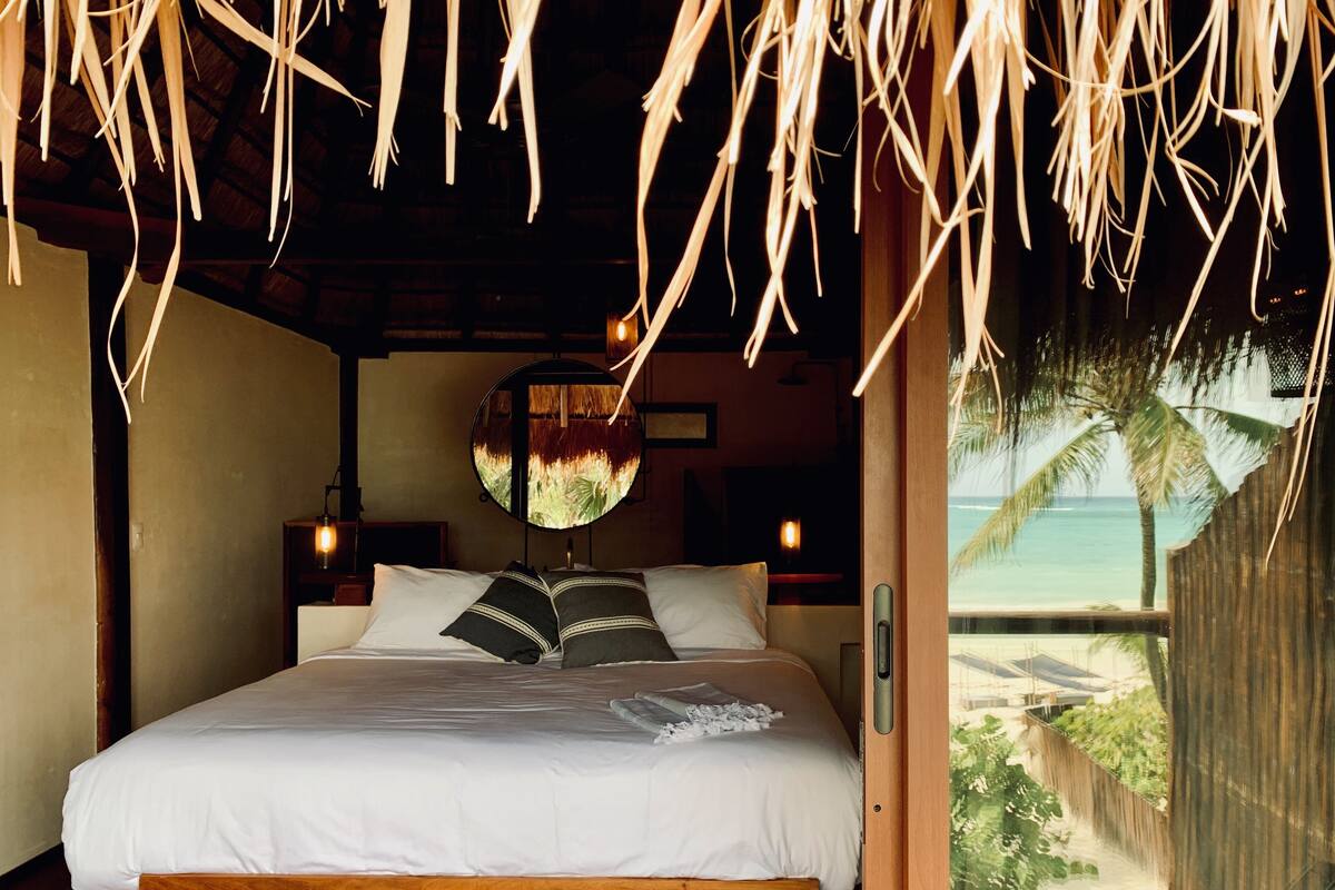 Quarto com cama de madeira, roupa de cama branca, espelho redondo e vista para a praia.