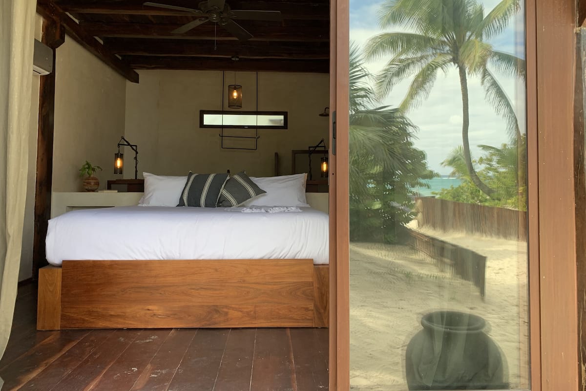 Quarto com cama de madeira, ventilador de teto e vista para uma praia com palmeiras.