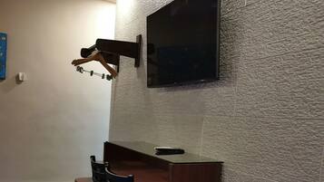Quarto com televisão na parede, uma cadeira de madeira e escrivaninha com controle remoto.