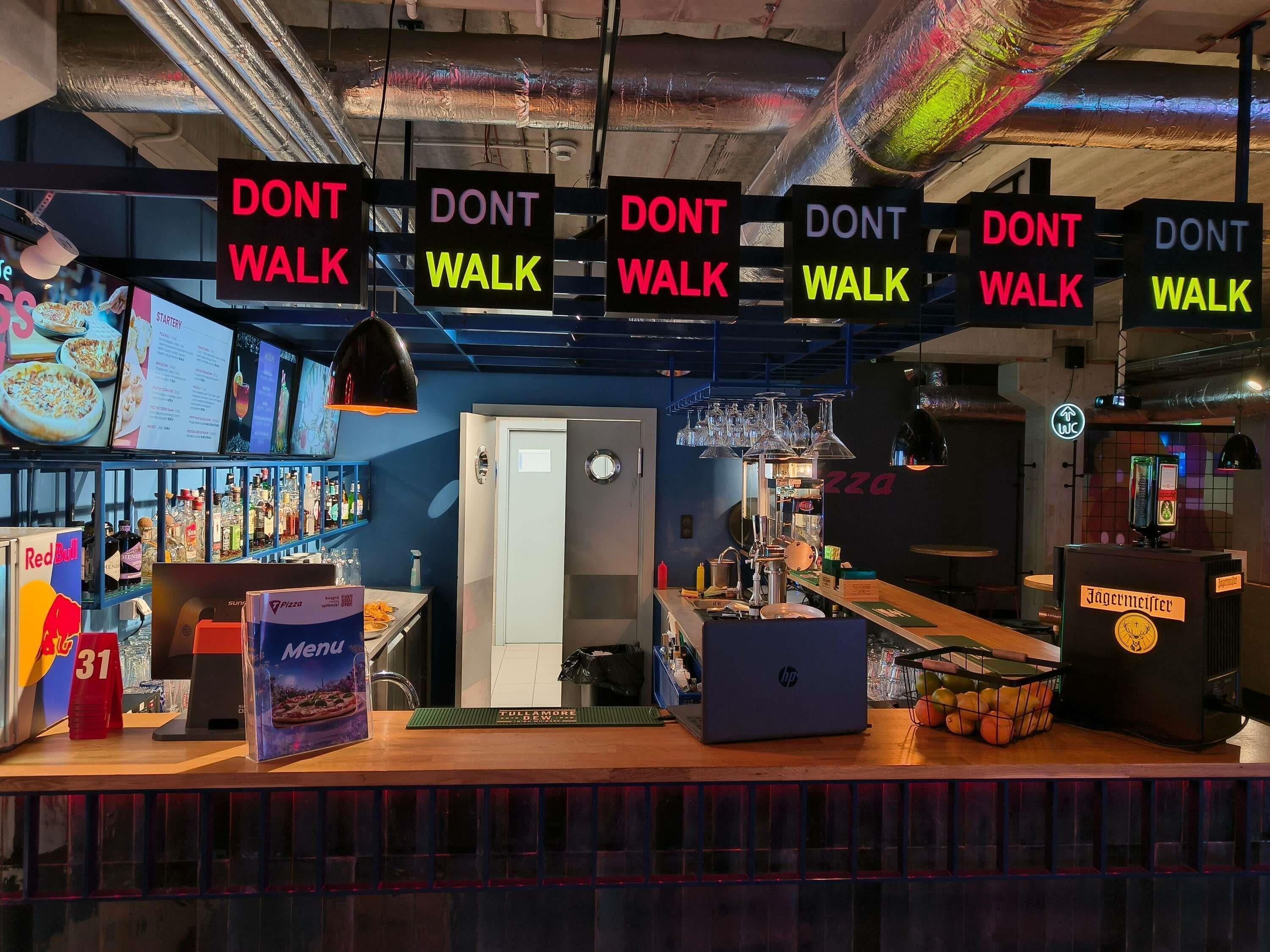 Uma área de bar com vários sinais de 'NÃO ANDE', um quadro de cardápio e uma geladeira com a inscrição 'Jagermeister'.