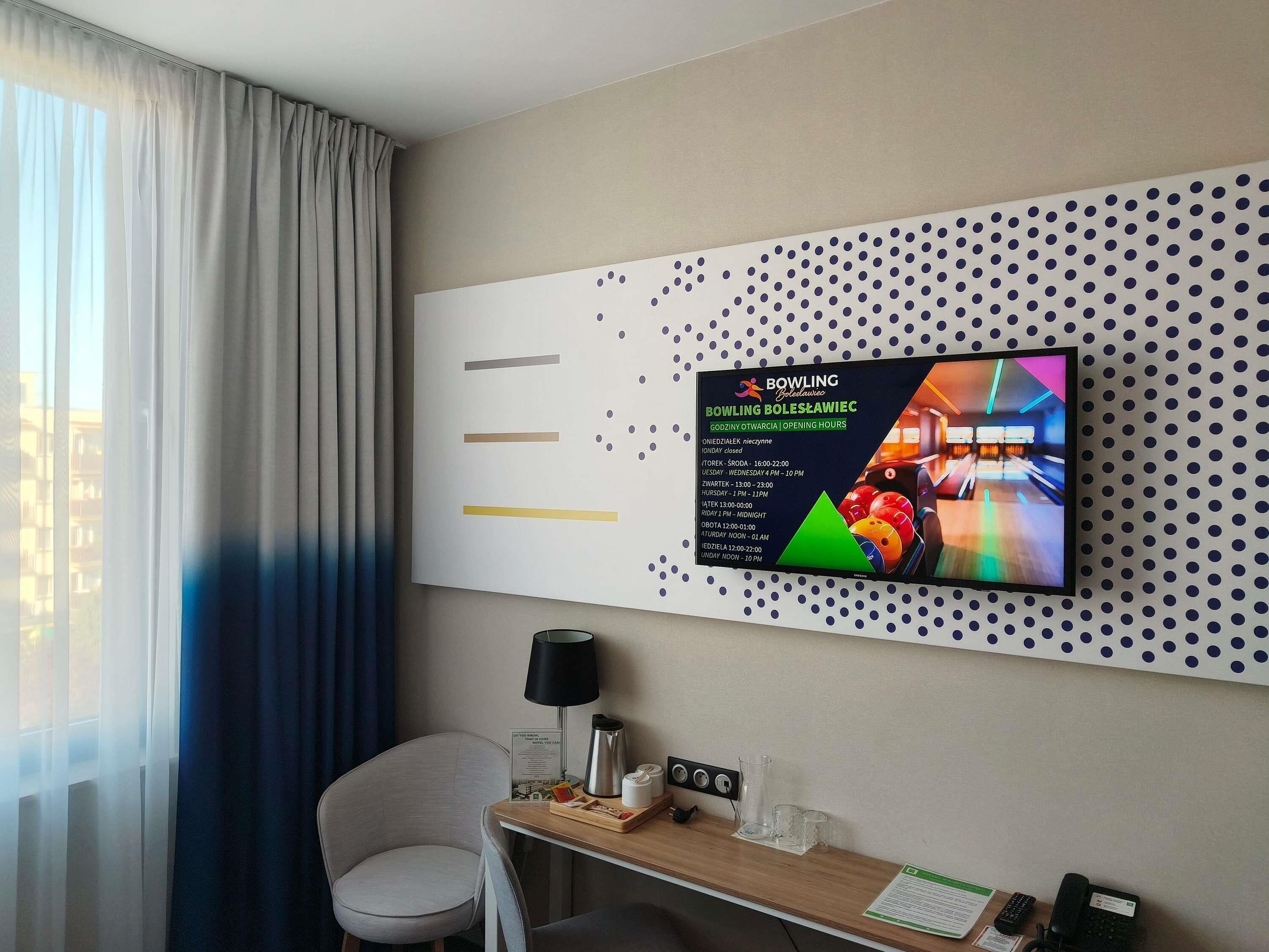 Quarto de hotel com televisão exibindo anúncio de boliche, escrivaninha com abajur, cadeira e vista para a cidade pela janela.
