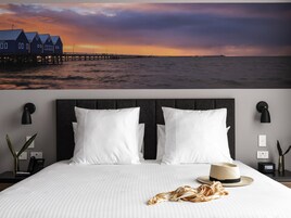 Quarto de hotel com uma cama, roupa de cama branca, um chapéu, um relógio e uma luminária na parede.