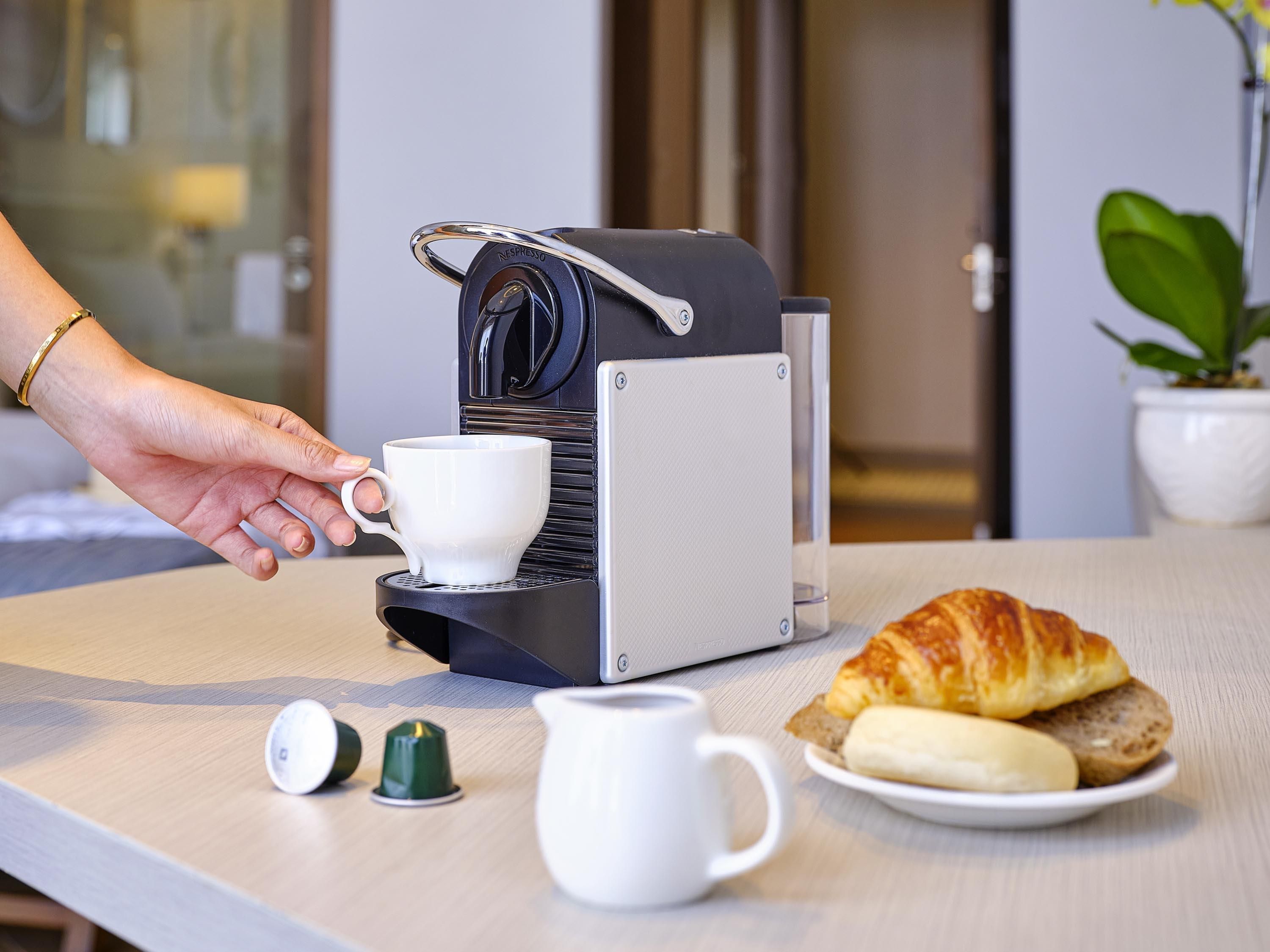 Uma máquina de café com porta-copos, um prato com croissant e pão, e uma pequena chávena branca sobre uma mesa.