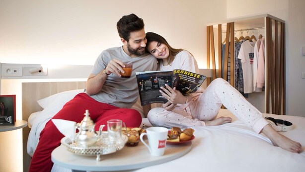 Um casal numa cama com uma revista, um tabuleiro com pastelaria e uma chávena.