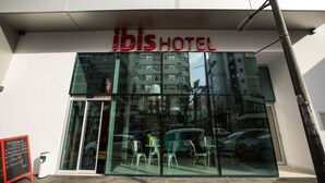 Exterior do hotel ibis com fachada de vidro, uma placa sobre a entrada e algumas cadeiras na parte externa.