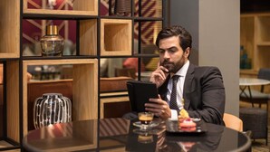 Um homem de terno sentado à mesa redonda com um laptop, um copo de bebida e um prato de comida. O ambiente inclui uma estante de madeira e um design de interiores moderno.