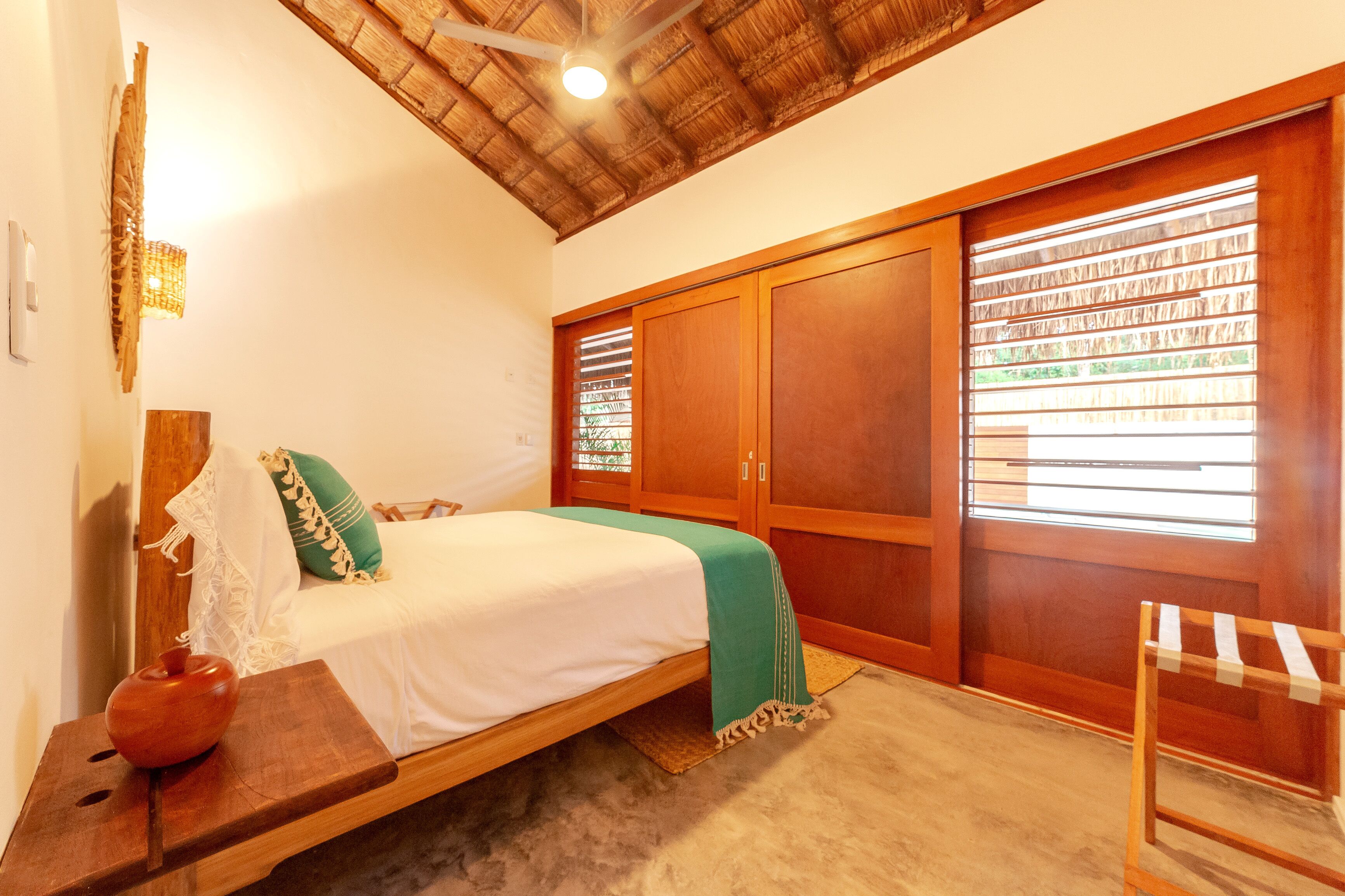 Quarto com cama, cabeceira de madeira, mesa de cabeceira com objeto decorativo, um banco de madeira, ventoinha de teto e uma janela grande com persianas.