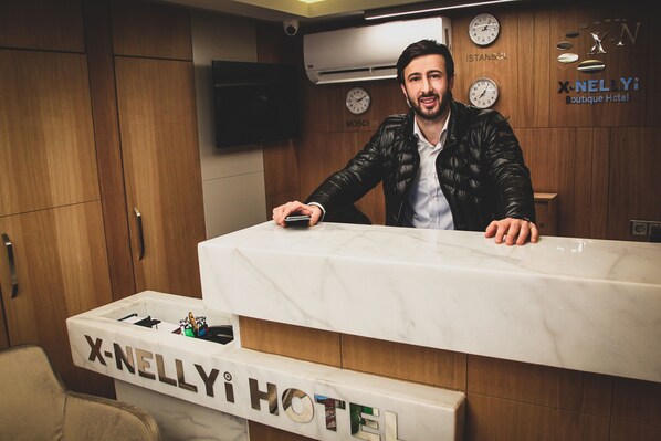Balcão de recepção de hotel com bancada de mármore, painéis de madeira, relógios e um letreiro com a inscrição 'X-NELLY iHOTEL'.