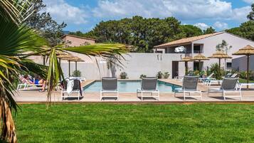 Área de piscina com espreguiçadeiras e guarda-sóis, cercada por vegetação e edifícios.
