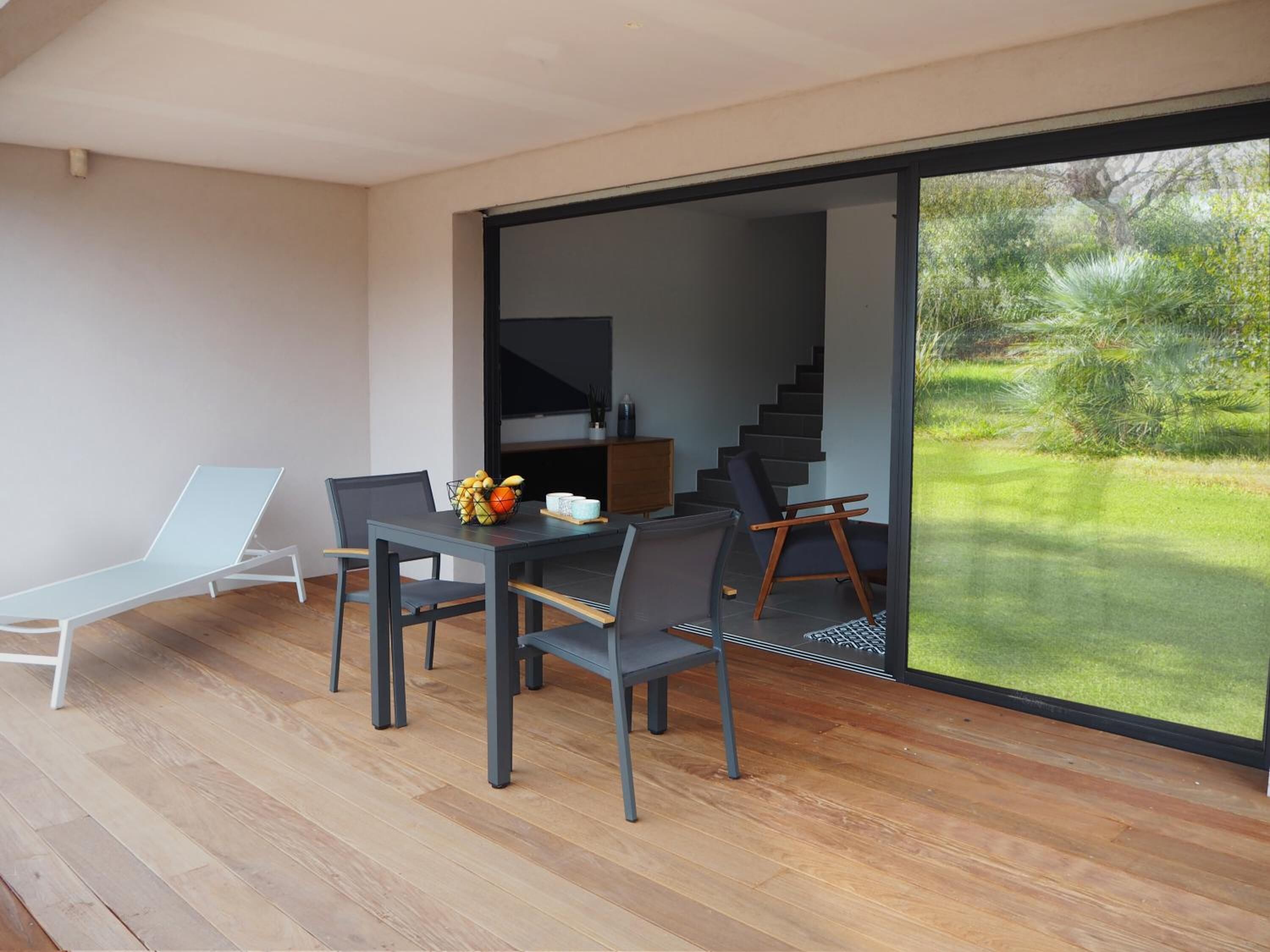 Uma área de refeições moderna ao ar livre, com mesa e cadeiras, uma poltrona de descanso e vista para um jardim através de uma porta de vidro.