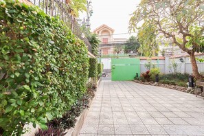 Uma trilha pavimentada que leva a um portão com grade verde, cercada por vegetação exuberante e uma árvore.