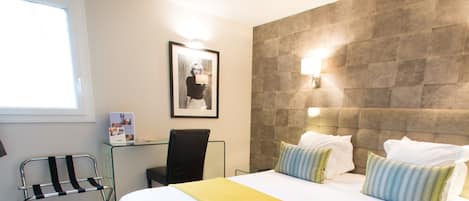Quarto de hotel com uma cama grande, duas luzes fixadas na parede, um quadro pendurado na parede, uma escrivaninha de vidro com uma cadeira e uma janela.