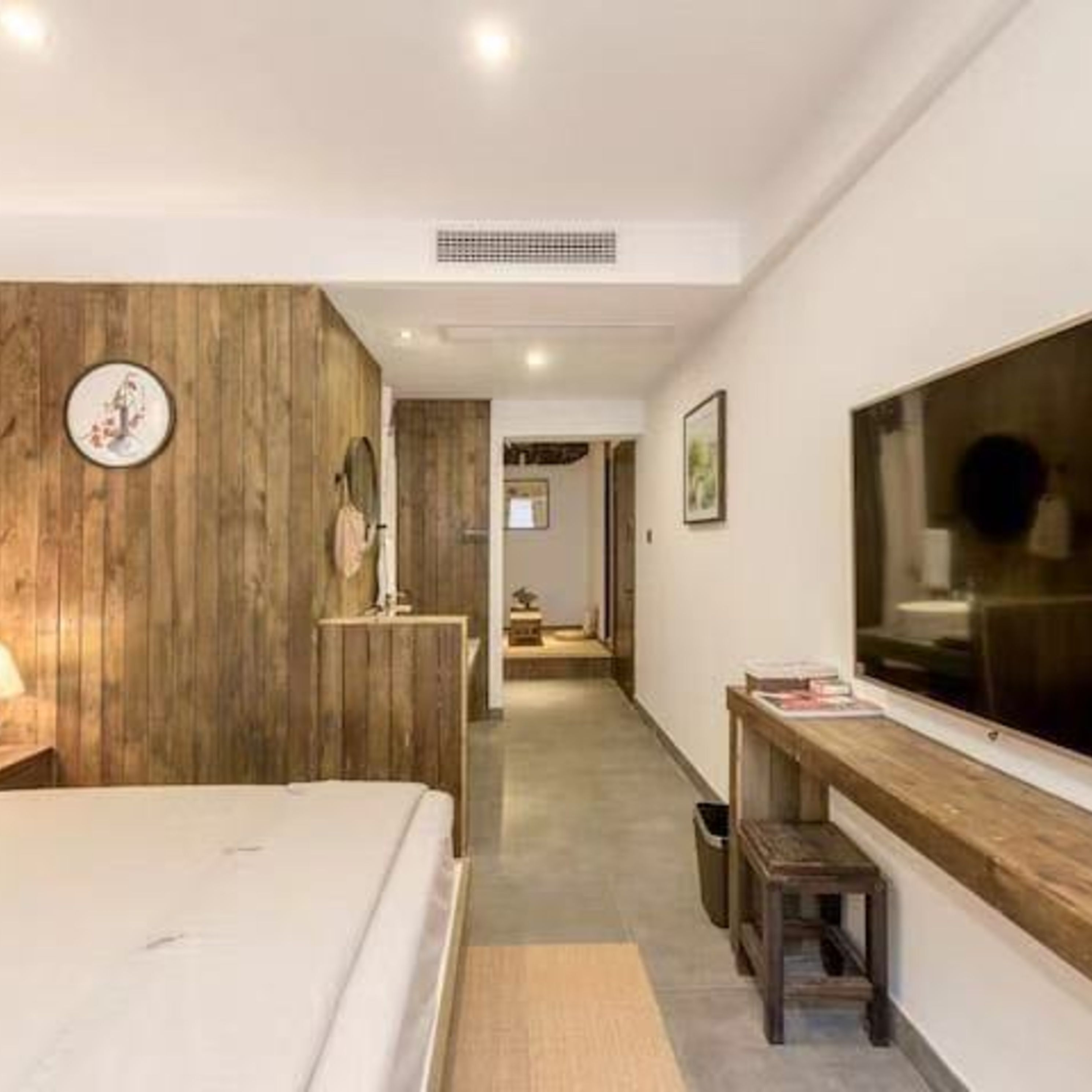 Quarto com parede em madeira decorativa, uma cama, mesa de cabeceira, espelho, televisão e um pequeno banquinho.