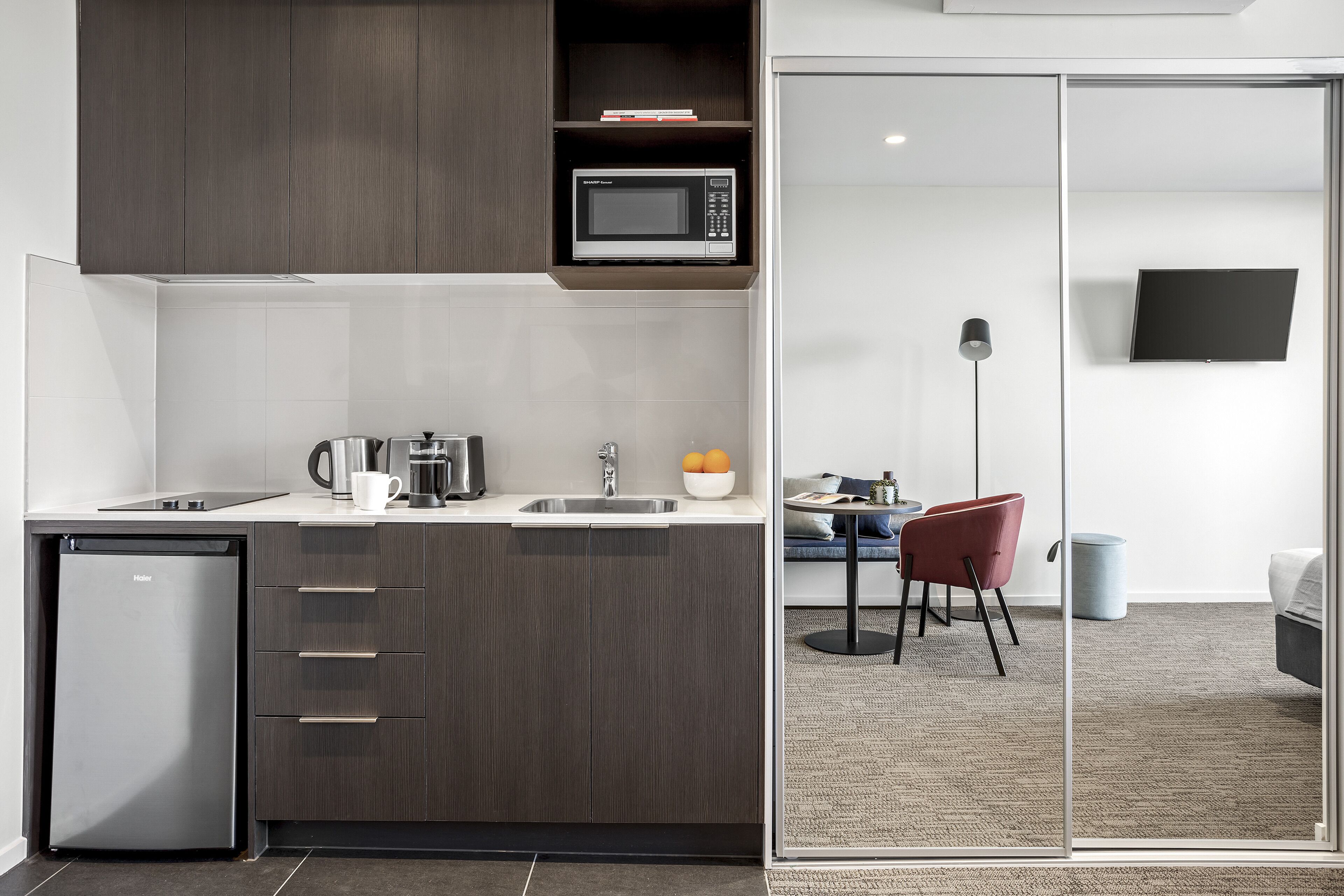 Cozinha moderna com micro-ondas embutido, cafeteira e pia. Um frigorífico compacto está à esquerda. Uma área de refeições com mesa e cadeiras é visível através de uma porta de vidro.