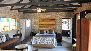 Quarto rústico com paredes de tijolos aparentes, teto de madeira, cama com roupa de cama floral, uma pequena mesa e uma janela com vitrais.
