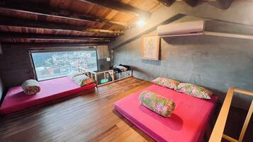 Quarto aconchegante com piso de madeira, duas camas com roupa de cama florida, uma janela com vista para edifícios e um ar condicionado fixado na parede.