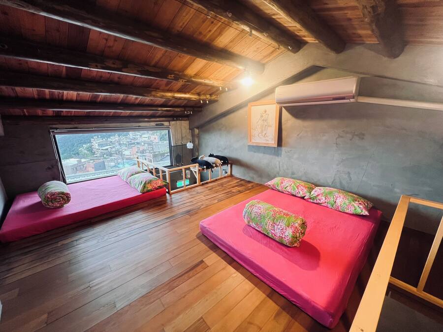 Quarto aconchegante com piso de madeira, duas camas com roupa de cama florida, uma janela com vista para edifícios e um ar condicionado fixado na parede.