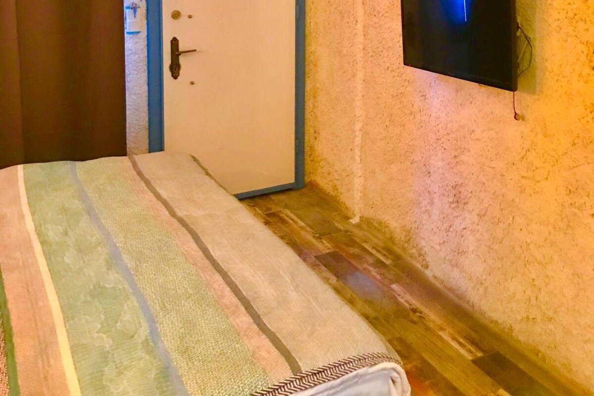 Quarto com cama, porta, televisão na parede e relógio.