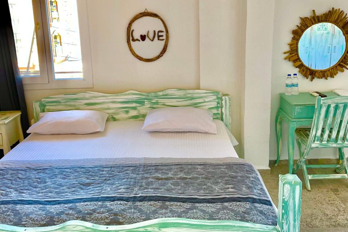 Um quarto com uma cama, ar condicionado fixado na parede, um adorno circular com a palavra 'LOVE', um espelho com moldura em forma de raio de sol e uma janela com vista para prédios.