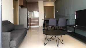 Uma sala de estar moderna com um sofá cinzento, uma mesa de centro redonda e uma TV sobre um móvel. Há também uma área de cozinha com armários de madeira e uma geladeira.