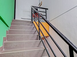 Uma escadaria com degraus cinzentos e corrimãos pretos, que leva a uma porta com um objeto vermelho no patamar.