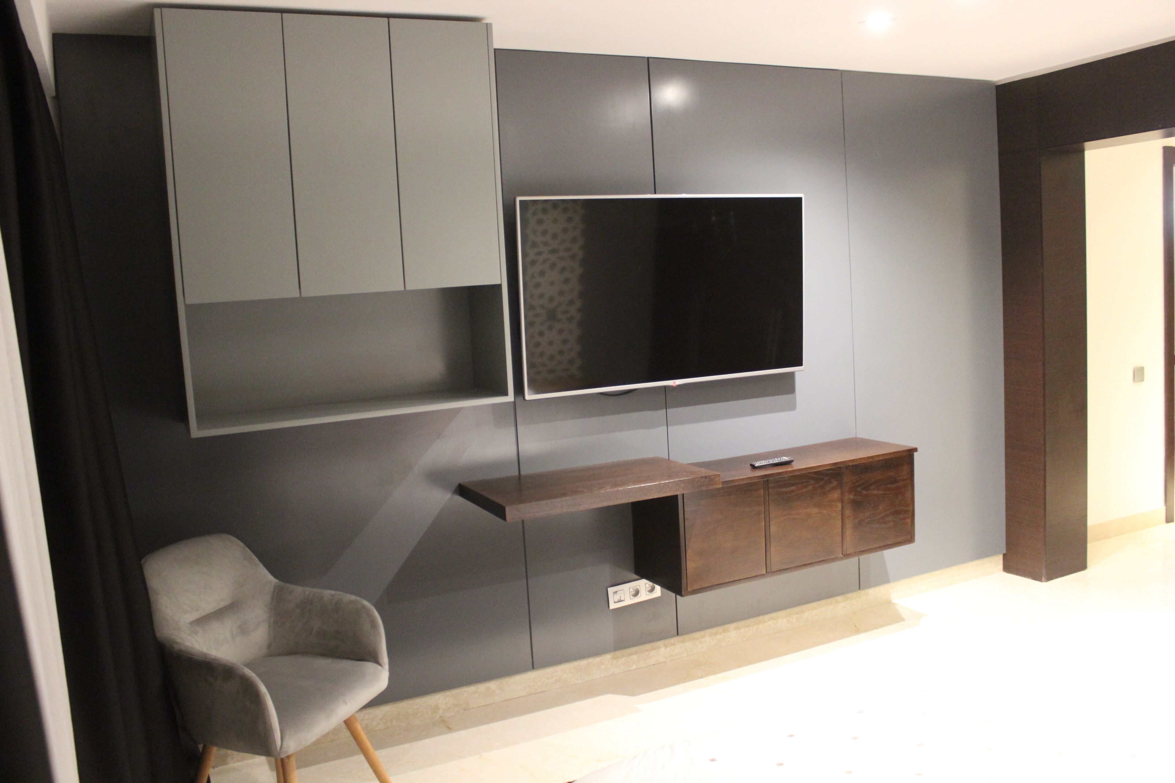Uma sala de estar moderna com uma televisão de tela plana presa a uma parede escura, um móvel de entretenimento de madeira abaixo e uma poltrona cinza.