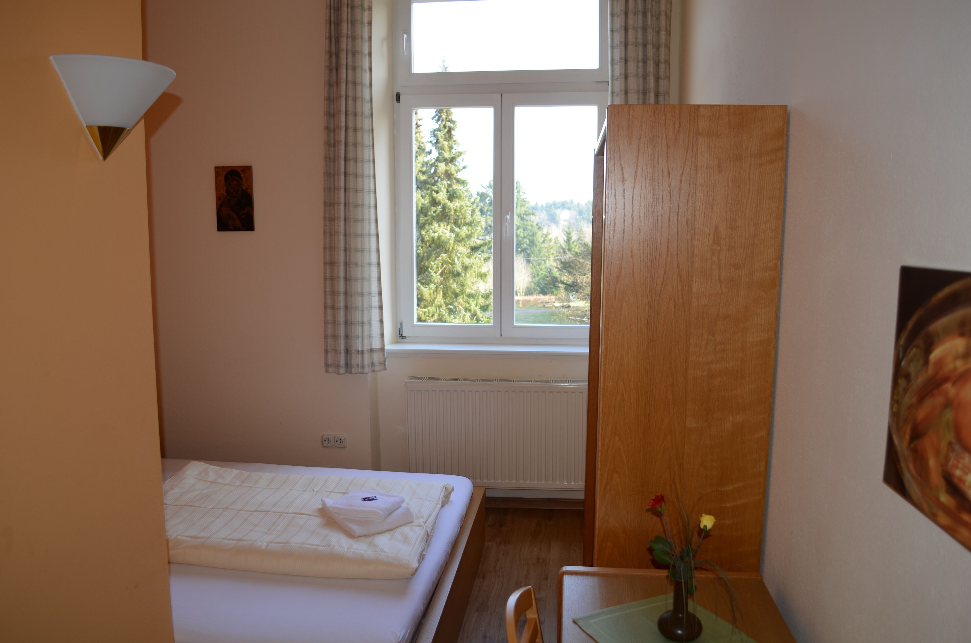 Quarto com cama, escrivaninha com cadeira, janela com cortinas, radiador e luminária fixada na parede.