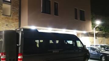Um parque de estacionamento com uma carrinha preta e um carro branco. Vê-se um edifício com várias janelas e um candeeiro sobre a carrinha.