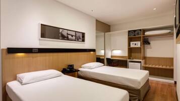 Quarto de hotel com duas camas de solteiro, uma escrivaninha com cadeira, um espelho e um guarda-roupa com prateleiras.