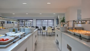 Uma área de buffet moderna e bem iluminada, com diversas estações de comida, incluindo frutas, saladas e doces. Há mesas e cadeiras dispostas para os hóspedes.
