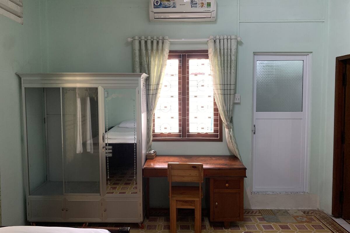 Quarto com cama, escrivaninha e cadeira, armário, janela com cortinas e porta.
