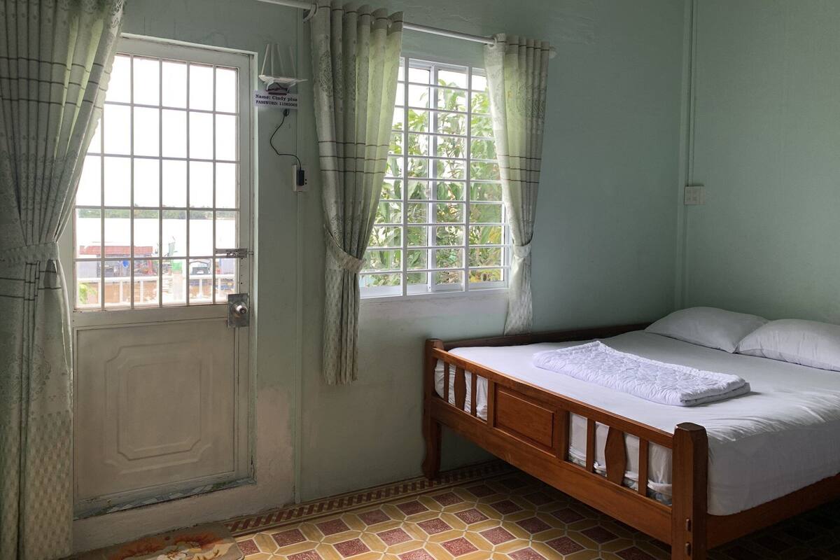 Um quarto com cama, janela com cortinas, porta e piso revestido com azulejos.