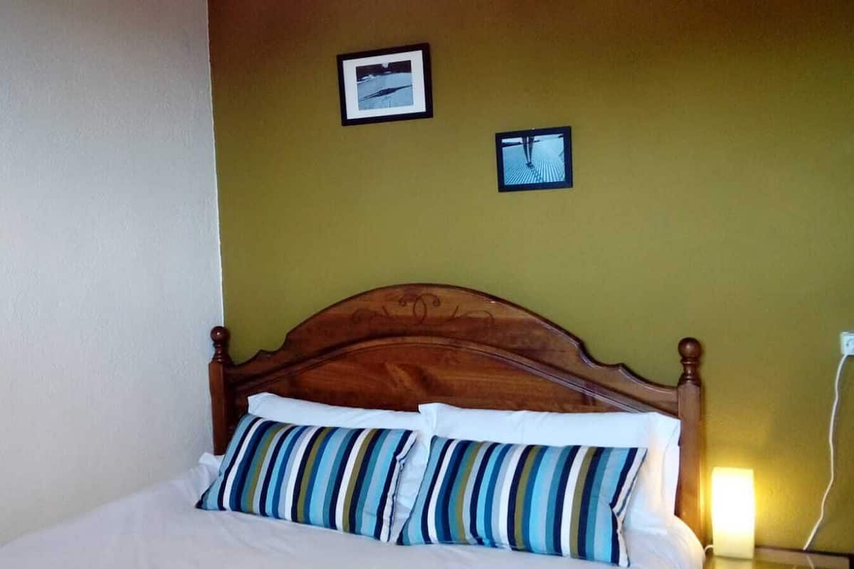 Uma cama com cabeceira de madeira, travesseiro listrado e parede lisa com duas molduras.