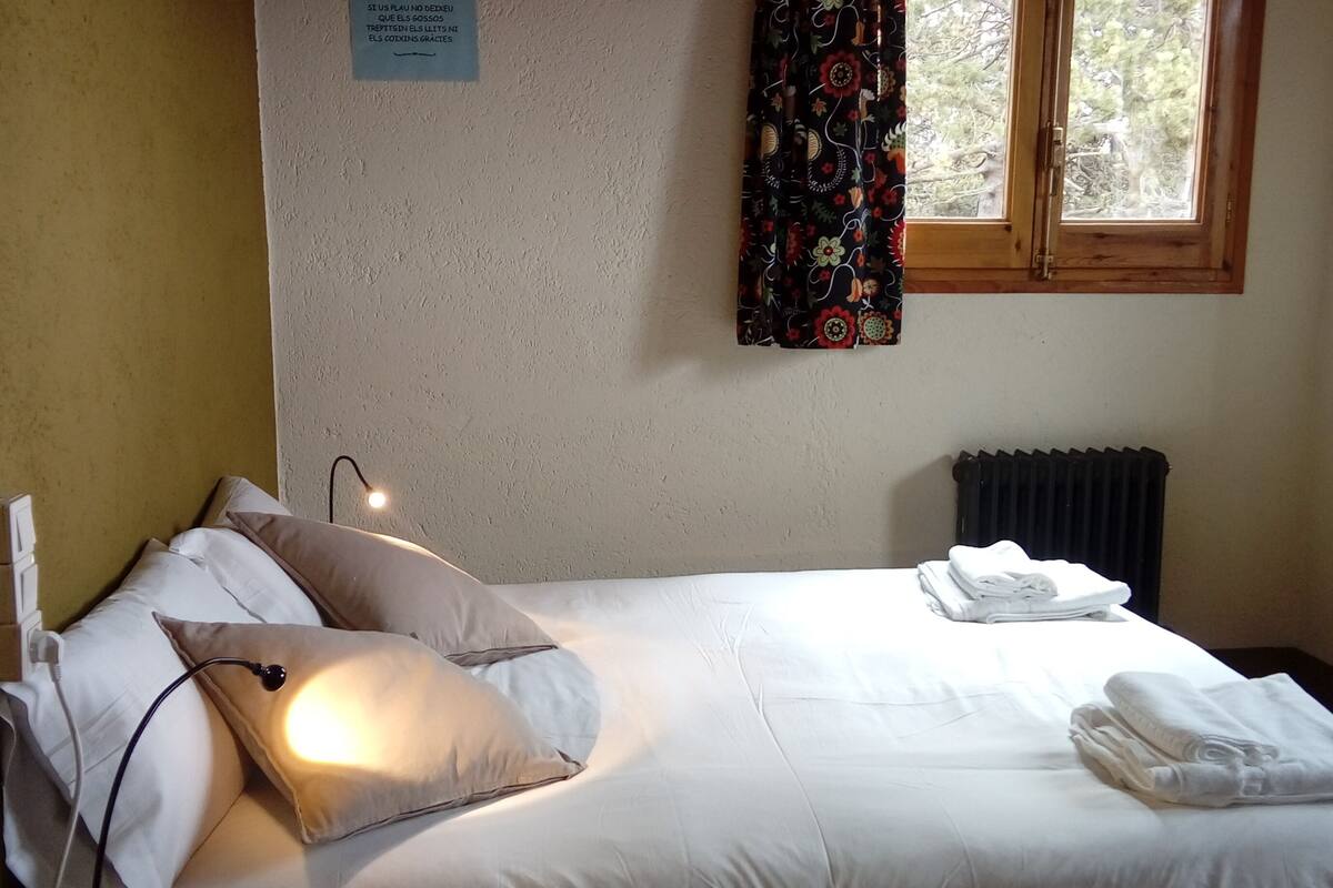 Quarto com cama, almofadas, abajur, radiador e janela com cortinas.