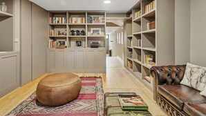 Uma sala de estar ampla, com um sofá de couro, um banquinho redondo e uma estante repleta de livros e objetos decorativos.