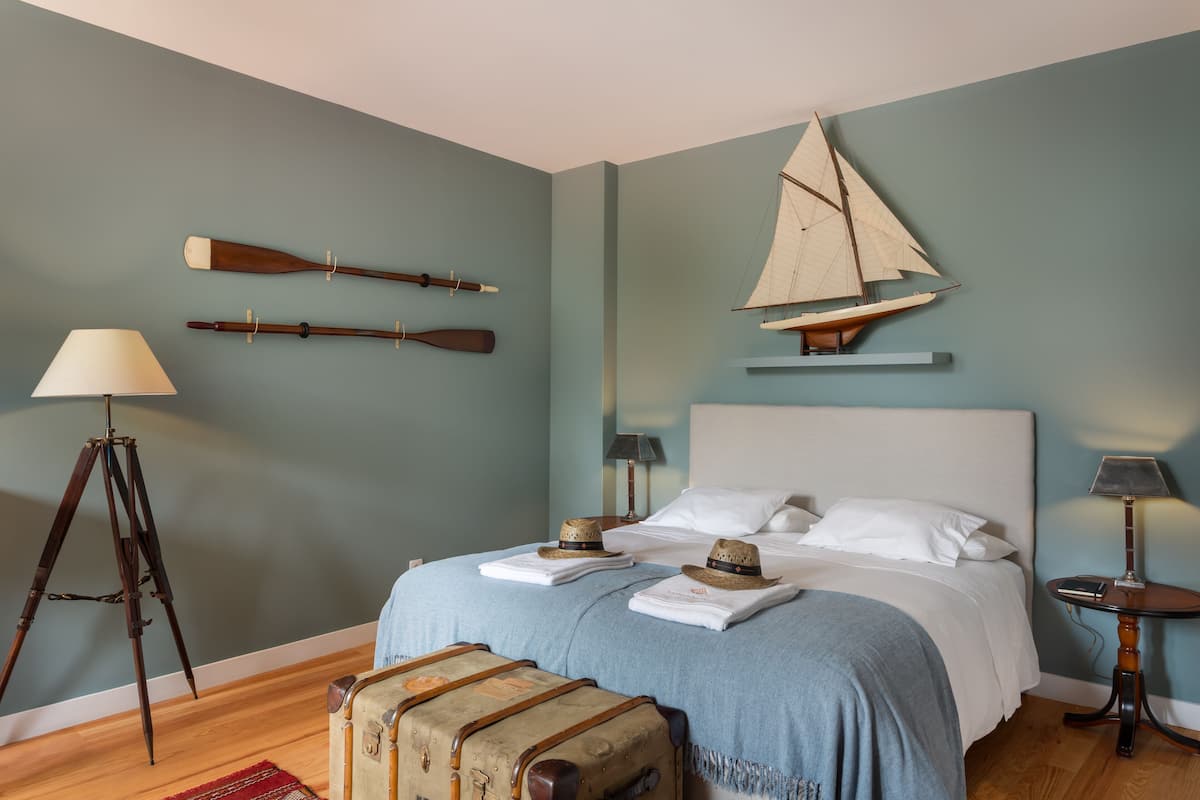 Quarto com cama, modelo de veleiro, mala antiga e abajur com tripé.