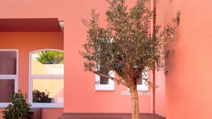 Uma pequena área externa com bancos e uma poltrona de descanso, uma planta em vaso e um edifício com fachada rosa e janelas.