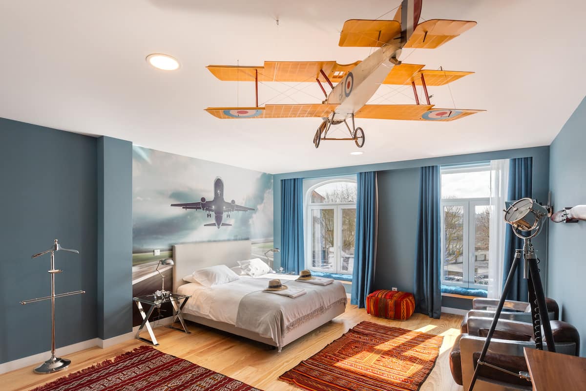 Quarto com cama, um modelo de avião pendurado, um tripé e janela com cortinas.