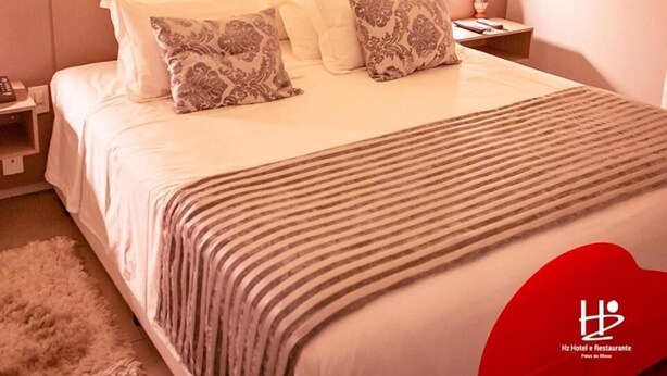 Quarto de hotel com cama bem arrumada, mesa de cabeceira com abajur e um logotipo vermelho na parede.