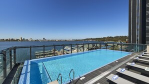 Uma piscina no terraço com água azul cristalina, horizonte urbano ao fundo e espreguiçadeiras com toalhas.