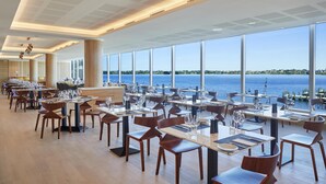 Um restaurante espaçoso com mesas preparadas para refeições, grandes janelas com vista para o mar e um design de teto moderno.