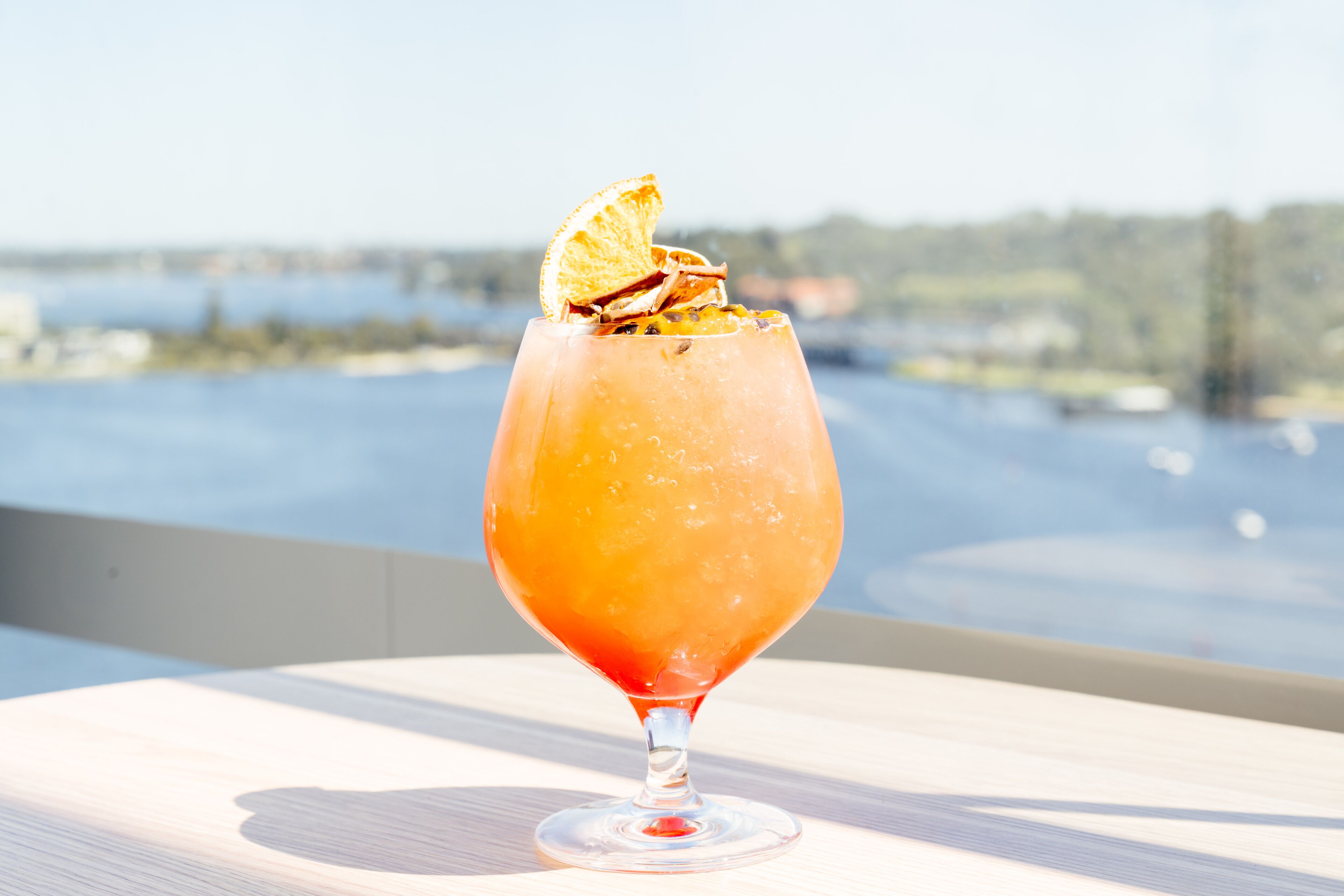 Um copo de cocktail com uma fatia de laranja na borda, apoiado em uma superfície de madeira, com vista para um corpo de água e edifícios ao fundo.