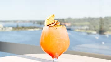 Um copo de cocktail com uma fatia de laranja na borda, apoiado em uma superfície de madeira, com vista para um corpo de água e edifícios ao fundo.