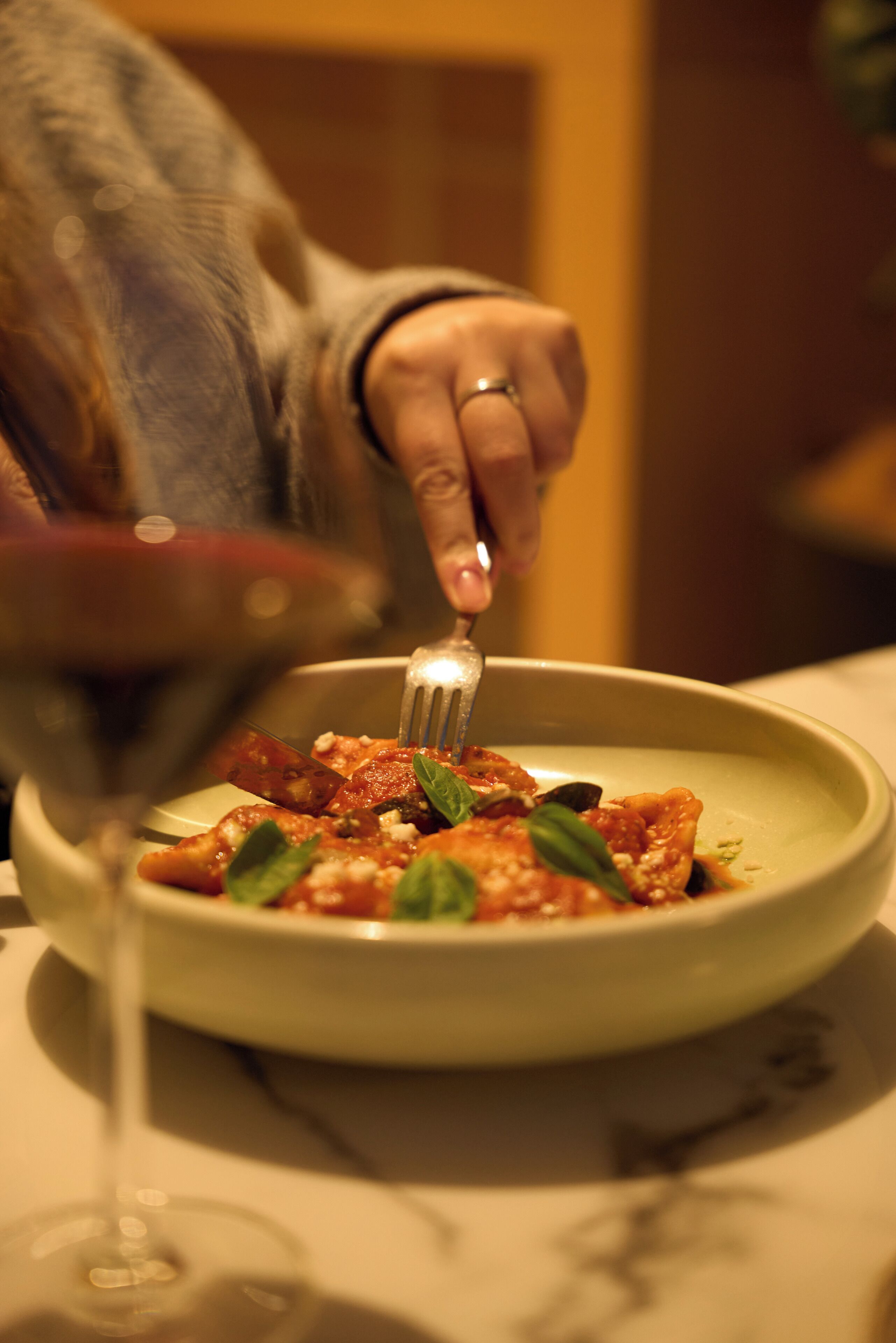 Uma pessoa segurando um garfo sobre um prato de comida, com um copo de vinho tinto em primeiro plano.