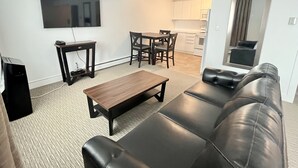 Uma sala de estar moderna com um sofá de couro, uma mesa de centro de madeira, uma TV fixada na parede e uma área de jantar com cadeiras e mesa.