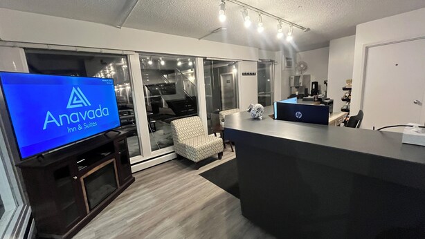 Uma sala de estar moderna com uma televisão exibindo 'Anavado Inn & Suites', uma janela grande com vista para uma escadaria e um balcão de recepção com um computador HP.