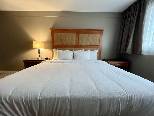 Quarto de hotel com uma cama grande, cabeceira de madeira, mesa de cabeceira com abajur e janela com cortinas.