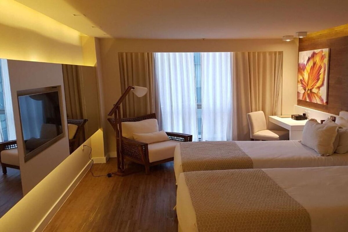 Quarto de hotel com duas camas, uma escrivaninha, uma cadeira, uma televisão e uma janela grande com cortinas.